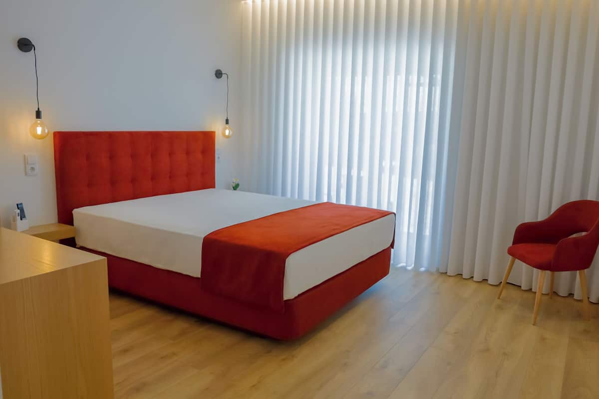 Quarto de hotel moderno com cama vermelha, piso de madeira, uma cadeira vermelha e uma janela grande com persianas verticais.
