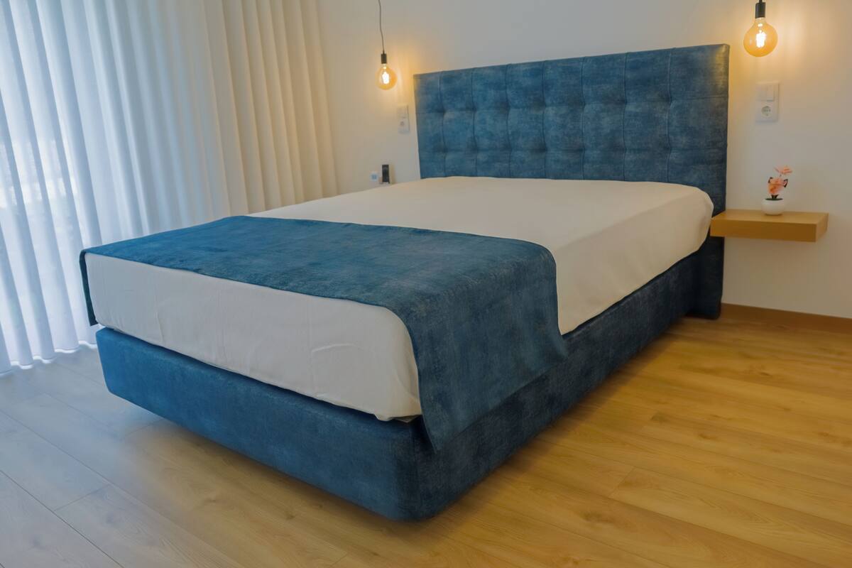 Quarto com cama, piso de madeira, cabeceira, mesas de cabeceira e janela com persianas.