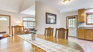Uma sala de jantar com mesa de madeira, cadeiras, um vaso com flores e vista para o exterior através de grandes janelas.