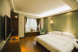 Quarto de hotel com cama, televisão, janela com cortinas, uma pequena mesa com uma planta e uma cadeira.
