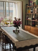 Uma mesa de jantar com toalha florida, um vaso com flores cor-de-rosa e um armário de madeira com garrafas e caixas.