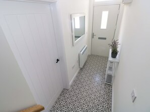 Um corredor estreito com ladrilhos estampados, uma porta branca, um radiador e um vaso de planta em uma prateleira.