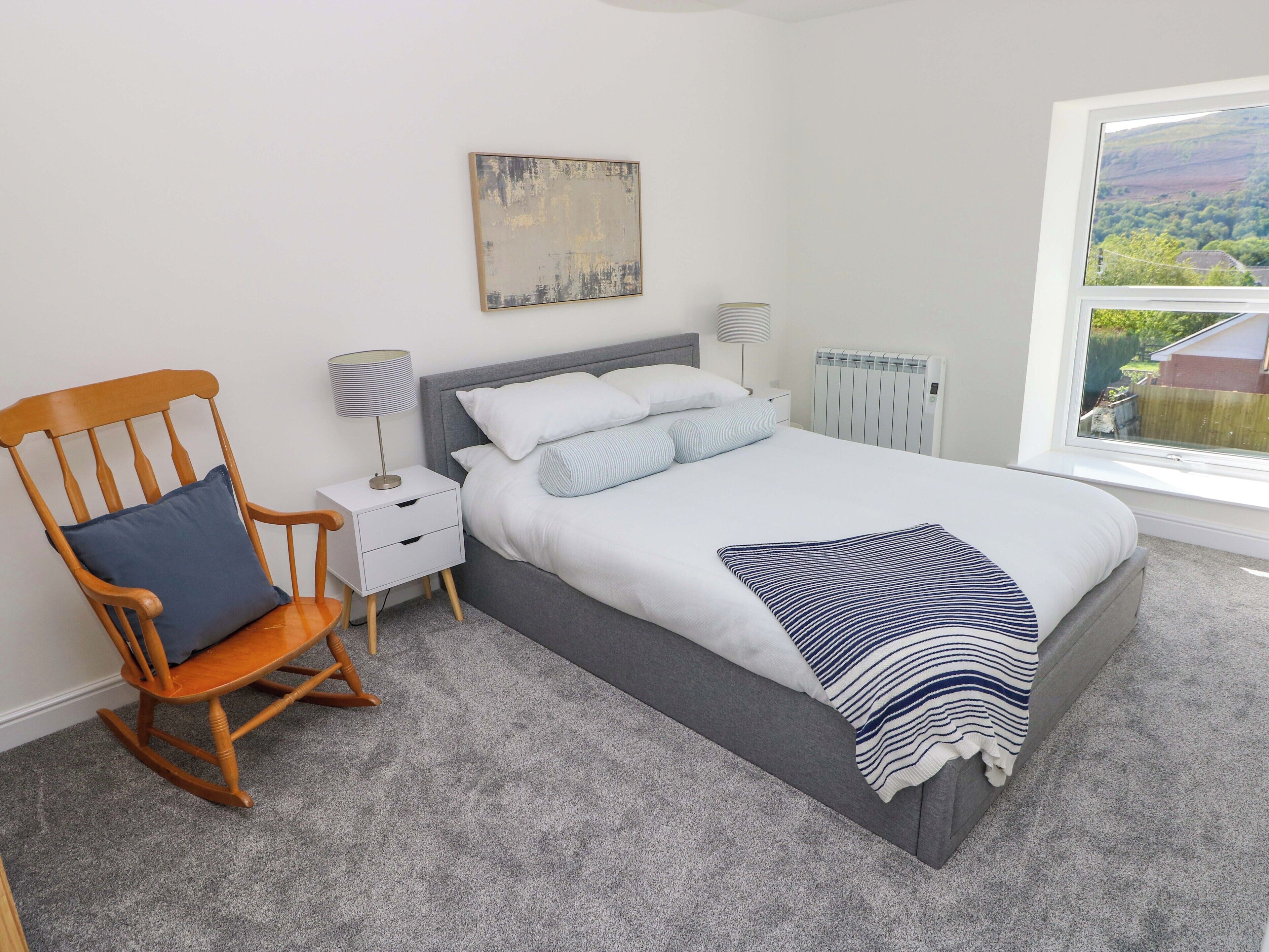 Quarto com cama, poltrona de balanço, mesa de cabeceira, luminária, radiador e janela com vista.