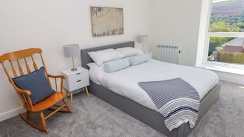 Quarto com cama, poltrona de balanço, mesa de cabeceira, luminária, radiador e janela com vista.