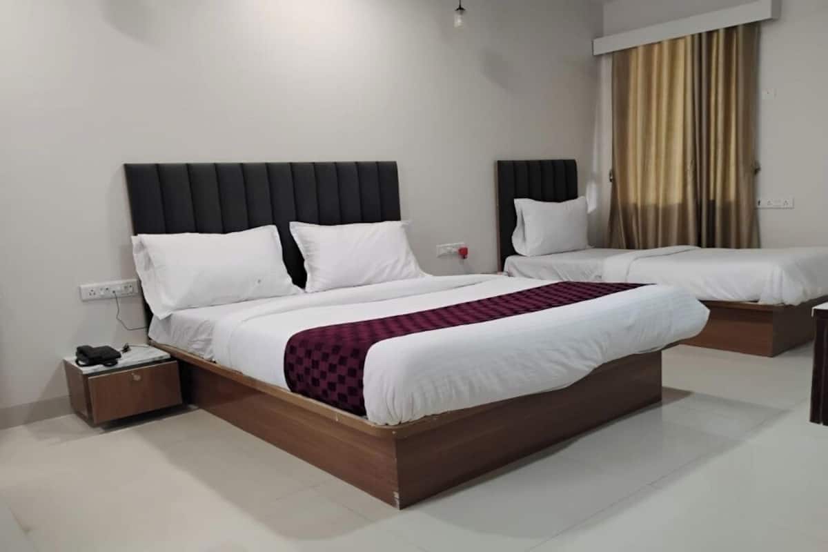 Cama de casal com cabeceira escura, mesa de cabeceira com telefone e carregador, e janela com cortinas.