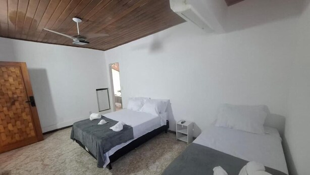 Quarto com duas camas, um ventilador de teto, uma porta de madeira e uma pequena mesa de cabeceira branca.