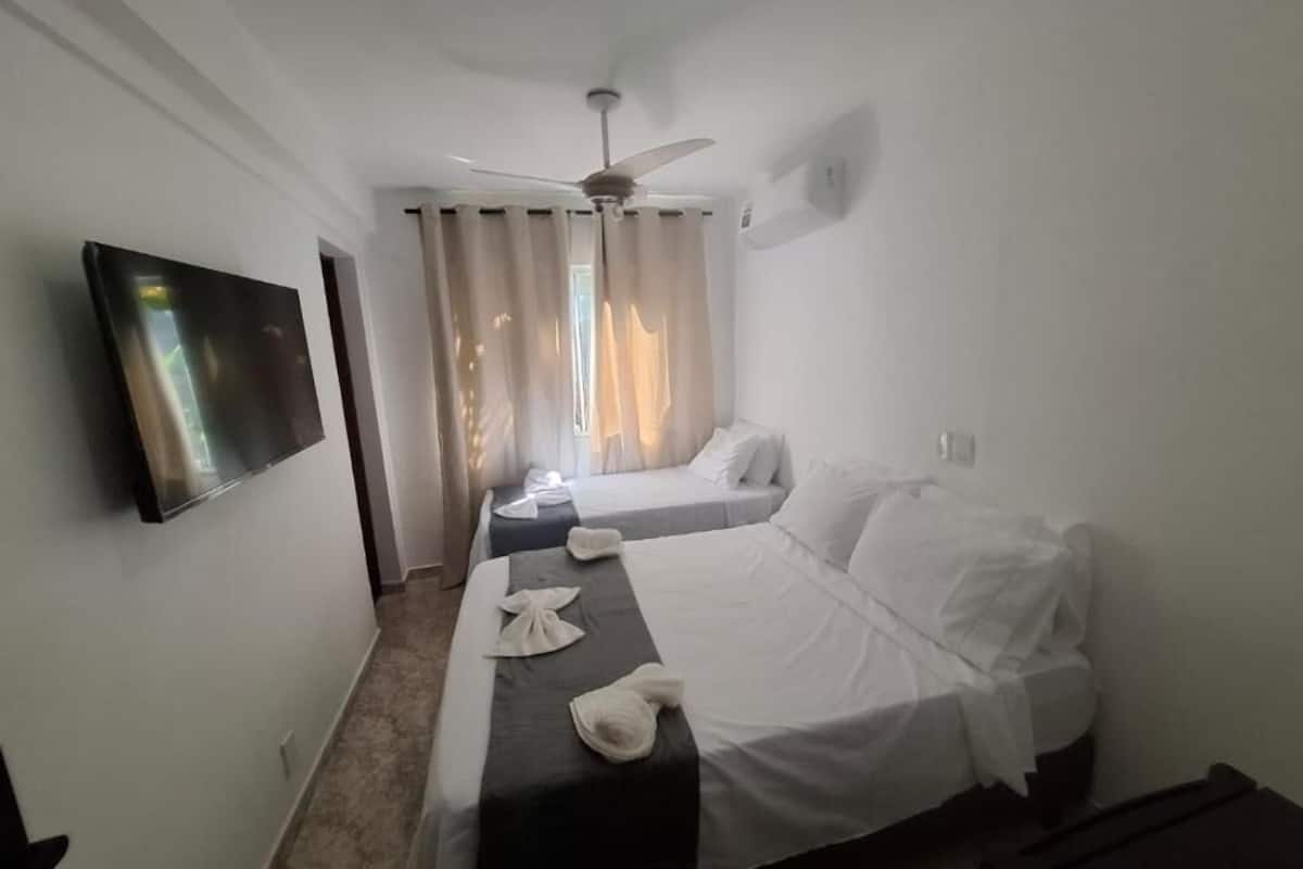 Quarto de hotel com cama, televisão fixada na parede, ventoinha no teto e janela com cortinas.