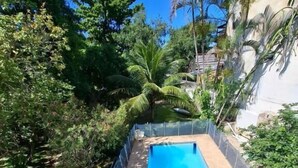 Piscina retangular rodeada por uma cerca de metal, com vegetação exuberante e um edifício ao fundo.