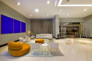 Um lobby moderno com um sofá curvo, cadeiras brancas, um banquinho laranja redondo e entrada de vidro.