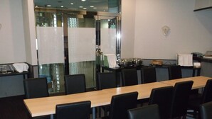 Uma sala de conferências com uma mesa comprida e cadeiras, uma área de cozinha com panelas e utensílios, e um painel de vidro.