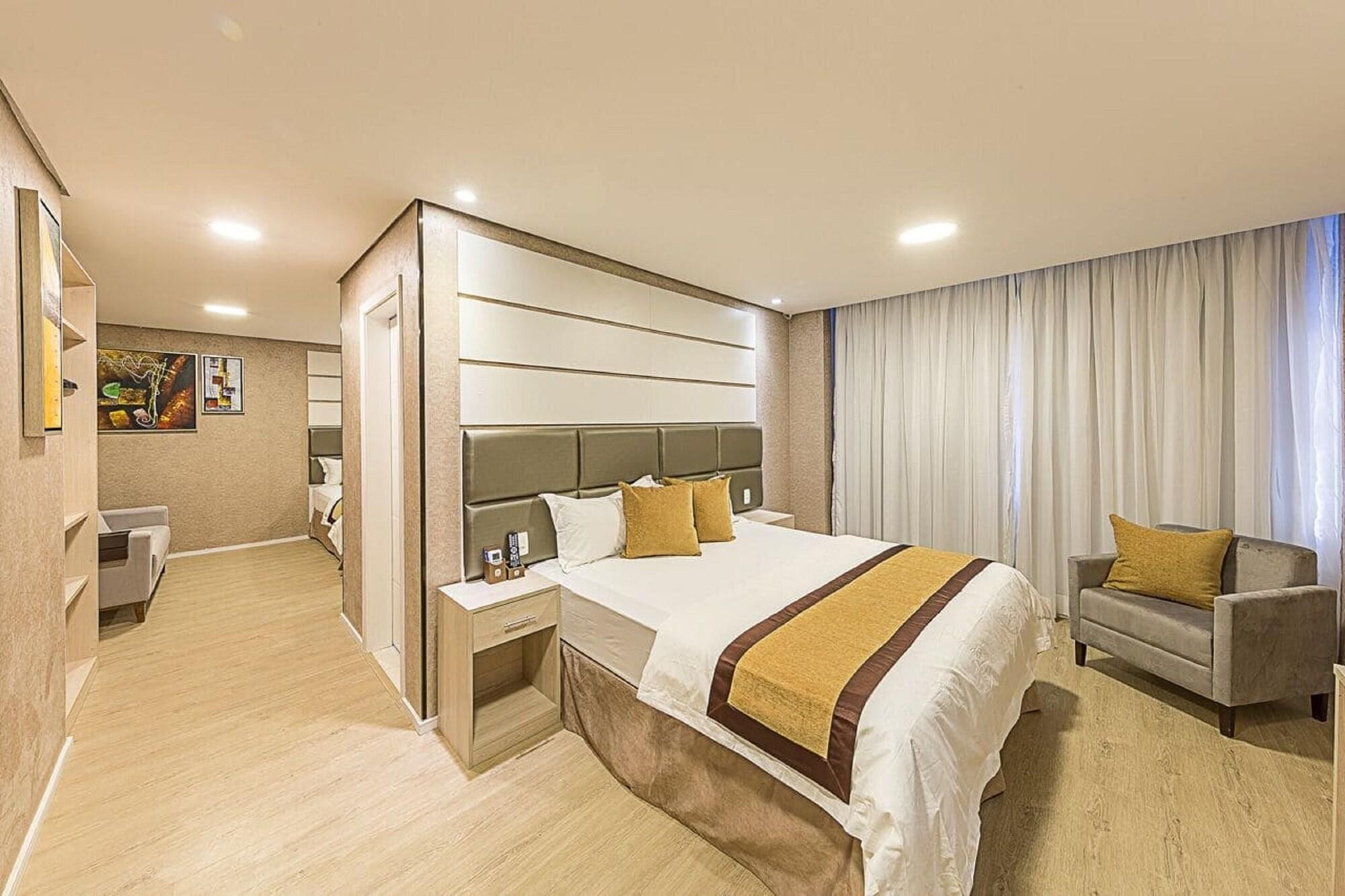 Quarto de hotel moderno com uma cama grande, mesas de cabeceira, uma escrivaninha, uma cadeira e vista para a cidade.