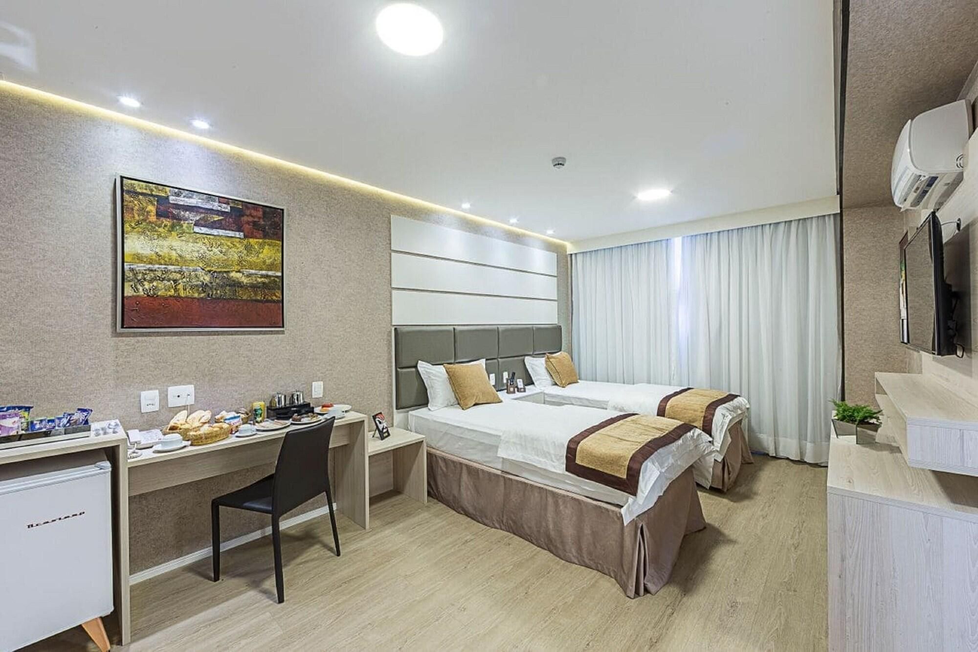 Quarto de hotel com duas camas, uma escrivaninha, uma cadeira, uma mesa de jantar e uma janela grande com cortinas.