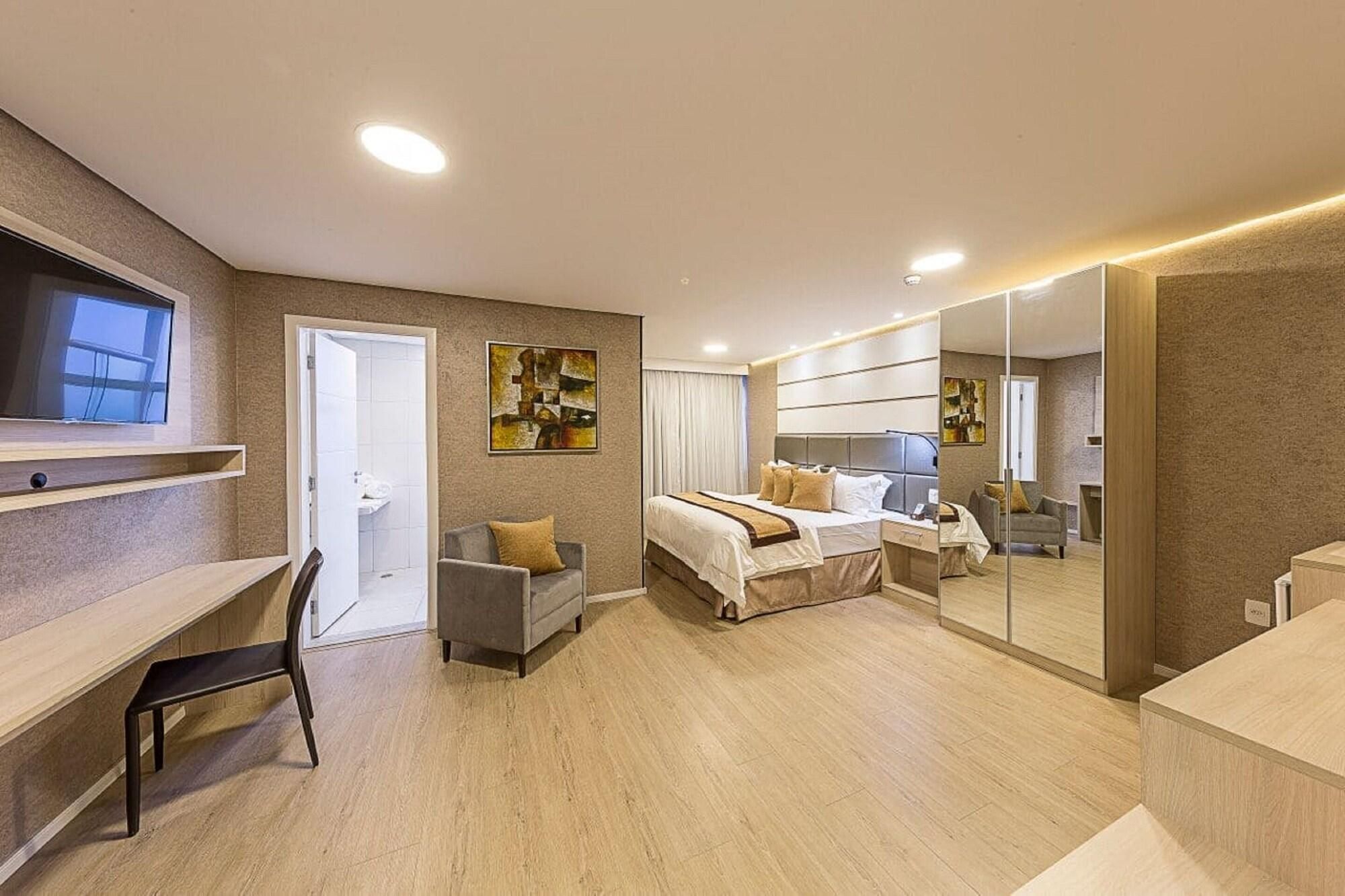 Quarto de hotel moderno com cama, escrivaninha, cadeira, espelho grande e obras de arte nas paredes.