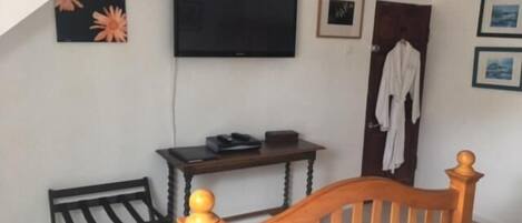 Quarto com cama, televisão fixada na parede, cadeira de madeira, mesa pequena e porta com roupão pendurado.
