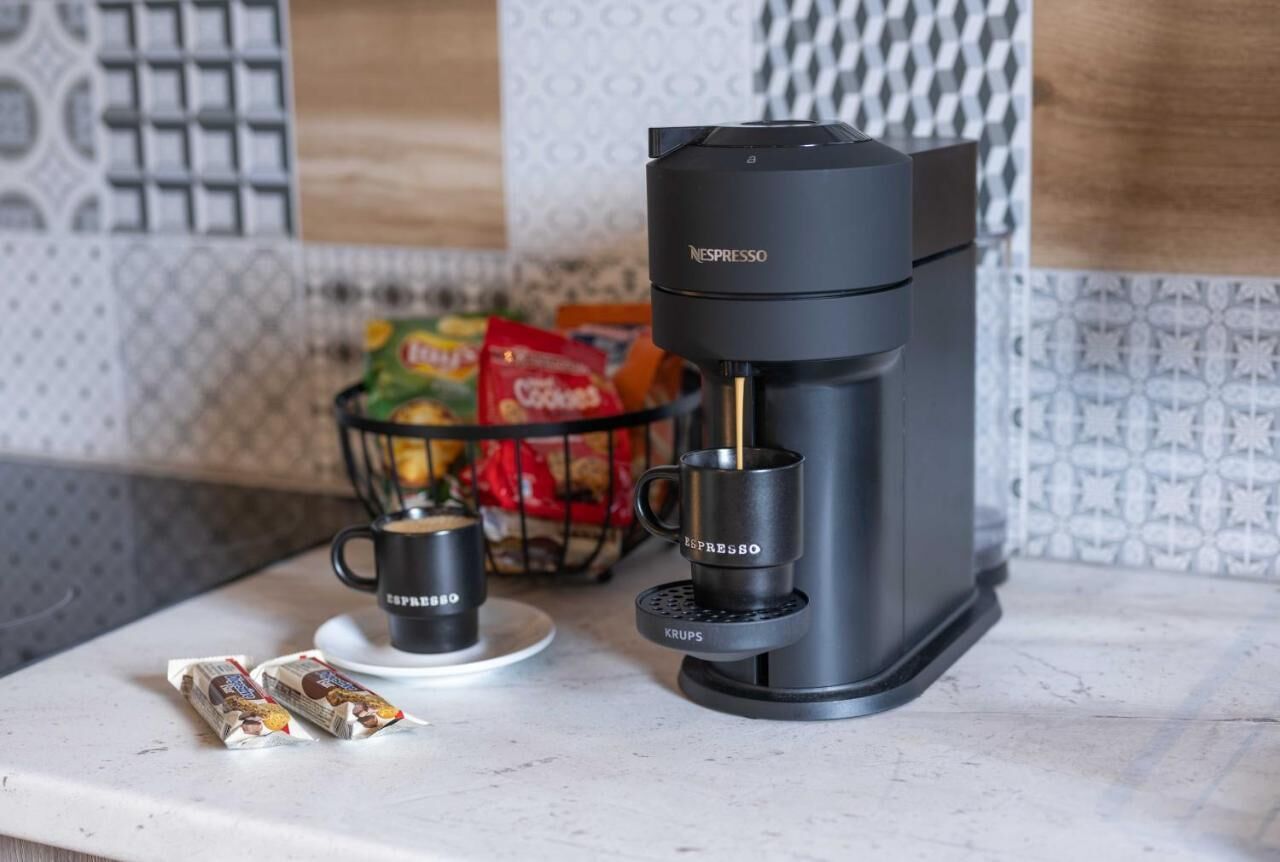 Uma cafeteira em uma bancada de cozinha, com uma xícara de café e dois sachês de creme para café.
