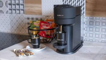Uma cafeteira em uma bancada de cozinha, com uma xícara de café e dois sachês de creme para café.