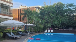 Piscina com espreguiçadeiras e guarda-sóis, rodeada por vegetação e um edifício ao fundo.