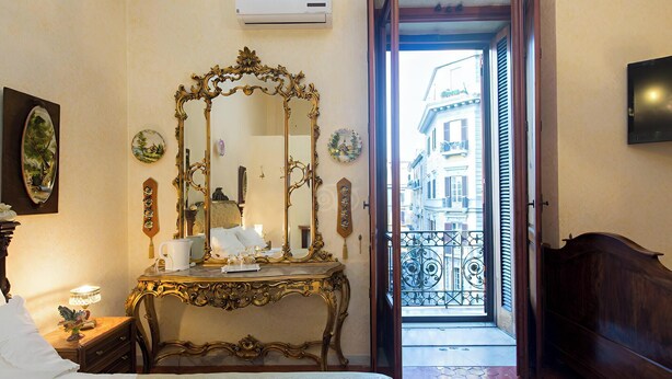 Um quarto com um espelho grande e ornamentado, uma pequena mesa com abajur e uma cama.