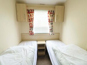 Acomodação pequena, com um quarto e duas camas de solteiro, cabeceira de madeira, janela com persianas e cortinas.