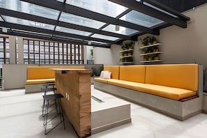 Interior moderno com teto de vidro, balcão de recepção em madeira, sofá amarelo e área de bar.