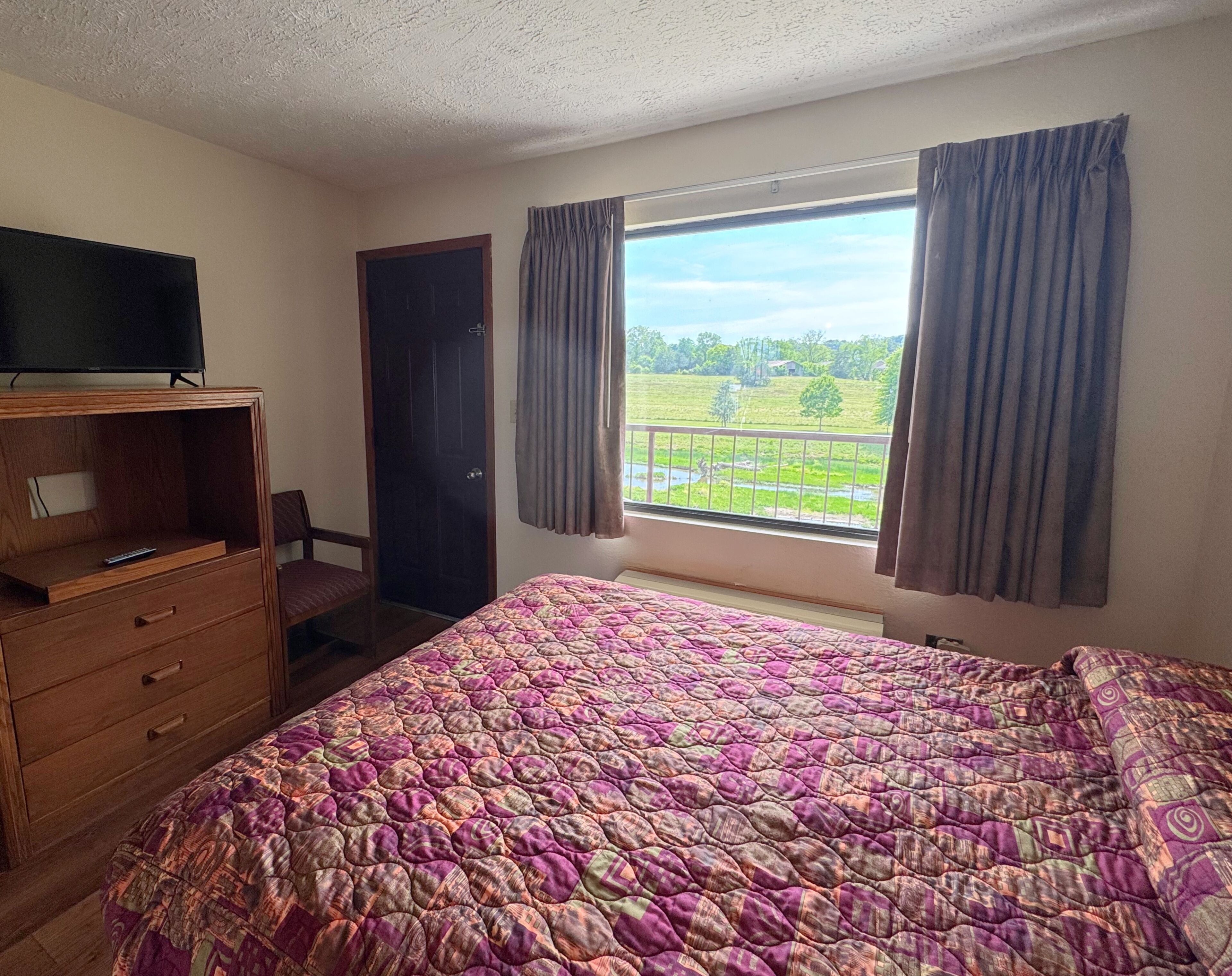 Quarto com cama, televisão sobre suporte de madeira, escrivaninha com gavetas, cadeira e janela com cortinas.