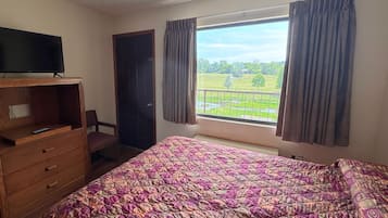 Quarto com cama, televisão sobre suporte de madeira, escrivaninha com gavetas, cadeira e janela com cortinas.