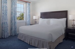 Quarto de hotel com uma cama grande, duas mesas de cabeceira, uma janela com cortinas e vista para palmeiras.