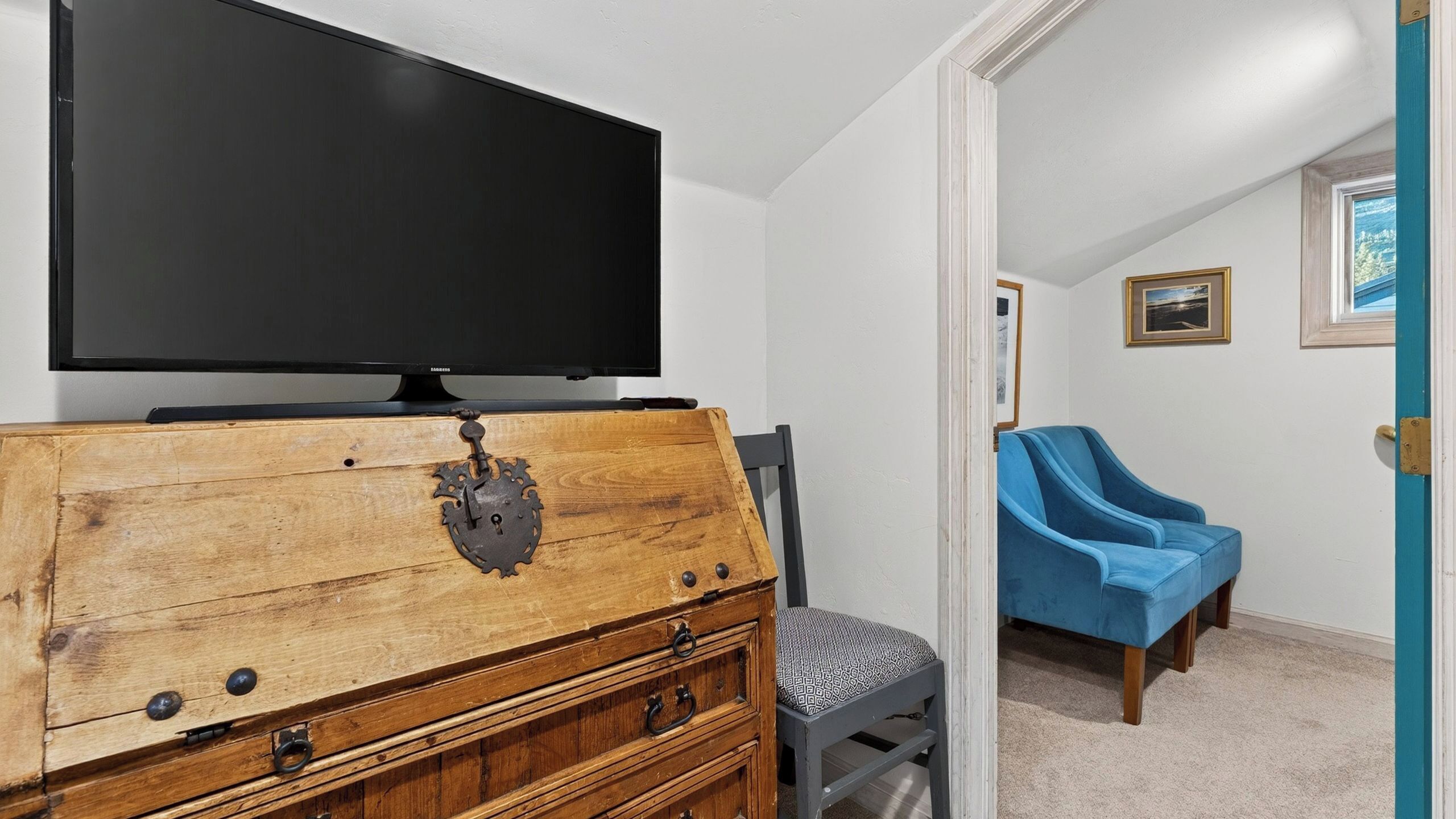 Quarto com cômoda de madeira, televisão montada acima, uma cadeira e uma cadeira azul com almofada.