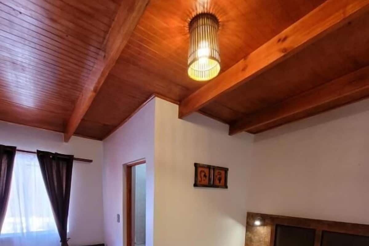 Quarto com cama, mesa e um banco. Há uma porta que dá para outro espaço.