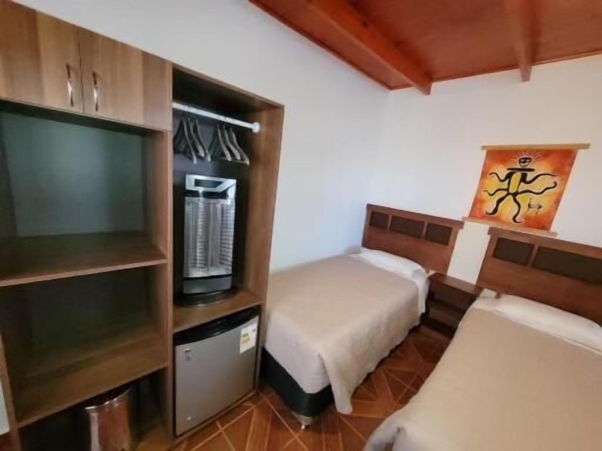 Quarto com duas camas, um guarda-roupa de madeira, um pequeno frigorífico e uma obra de arte na parede.