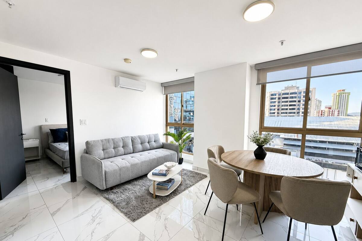 Uma sala de estar moderna com sofá, mesa de jantar, cadeiras, uma planta em vaso e vista para a cidade através de grandes janelas.