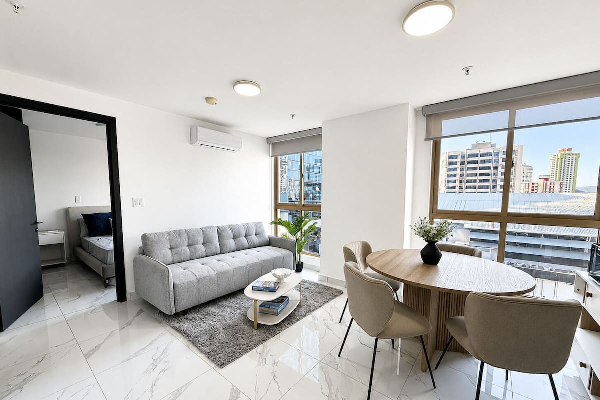 Uma sala de estar moderna com sofá, mesa de jantar, cadeiras, uma planta em vaso e vista para a cidade através de grandes janelas.