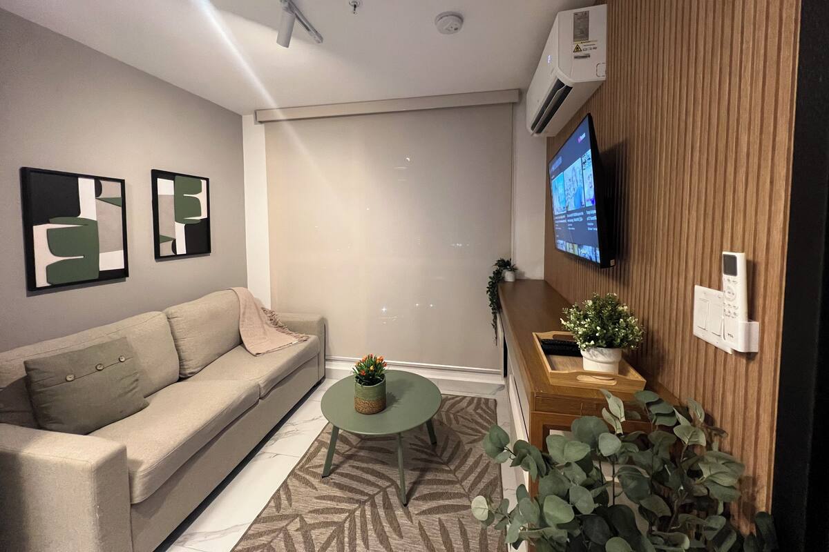 Uma sala de estar moderna com um sofá, uma mesinha redonda com uma planta, uma TV fixada na parede e um ar condicionado também fixado na parede.