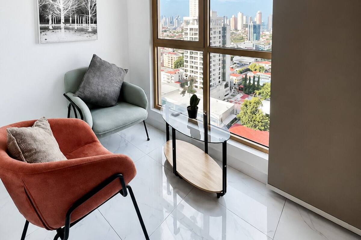 Sala de estar moderna com uma grande janela, vista da cidade, uma fotografia em preto e branco emoldurada e uma pequena mesa com uma planta.