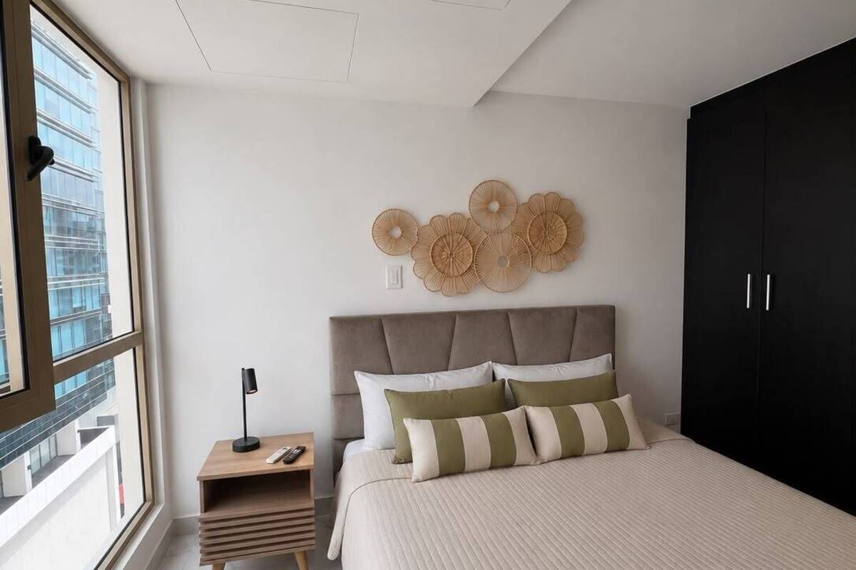 Um quarto moderno com uma cama grande, uma mesa de cabeceira, um candeeiro fixado na parede e vista para a cidade através de uma janela ampla.