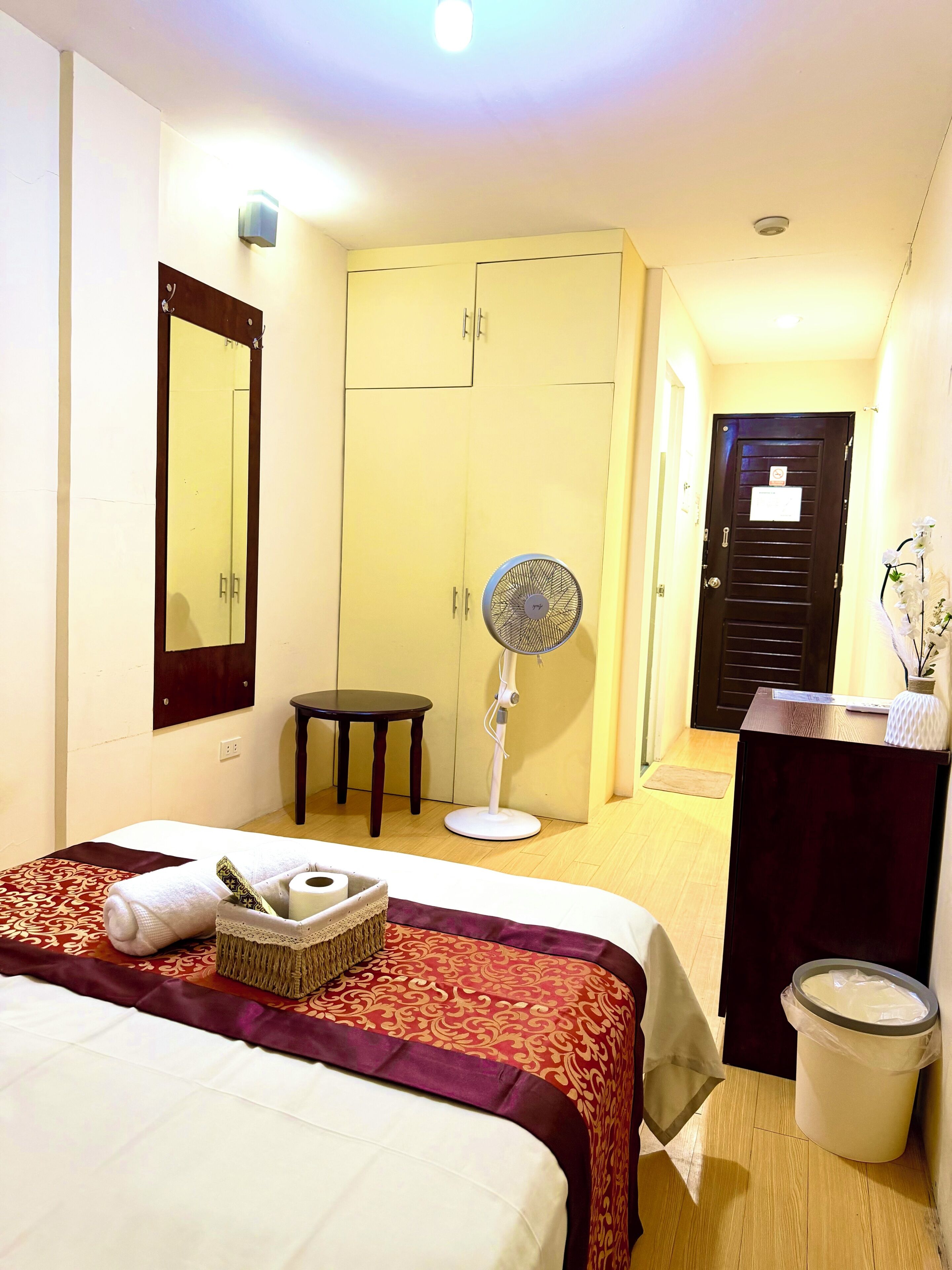 Quarto de hotel com cama, mesa de cabeceira, ventilador, guarda-roupa e lixeira.