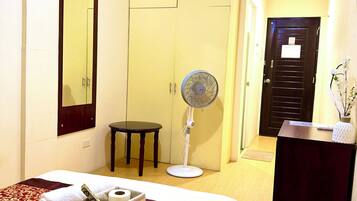 Quarto de hotel com cama, mesa de cabeceira, ventilador, guarda-roupa e lixeira.