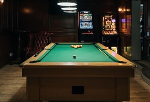 Uma mesa de bilhar com bolas, uma cadeira e duas máquinas de jogo iluminadas em um ambiente com pouca luz.