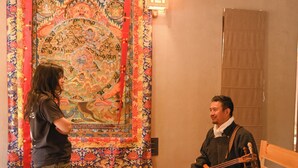 Um quarto com teto de madeira, uma tapeçaria grande, um tapete estampado e uma pessoa tocando um instrumento de cordas.