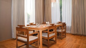 Uma sala de jantar com mesa e cadeiras de madeira, janelas amplas com cortinas e um lustre.