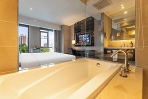 Quarto de hotel moderno com uma banheira grande, uma TV fixada na parede e uma varanda com vista para a paisagem urbana.