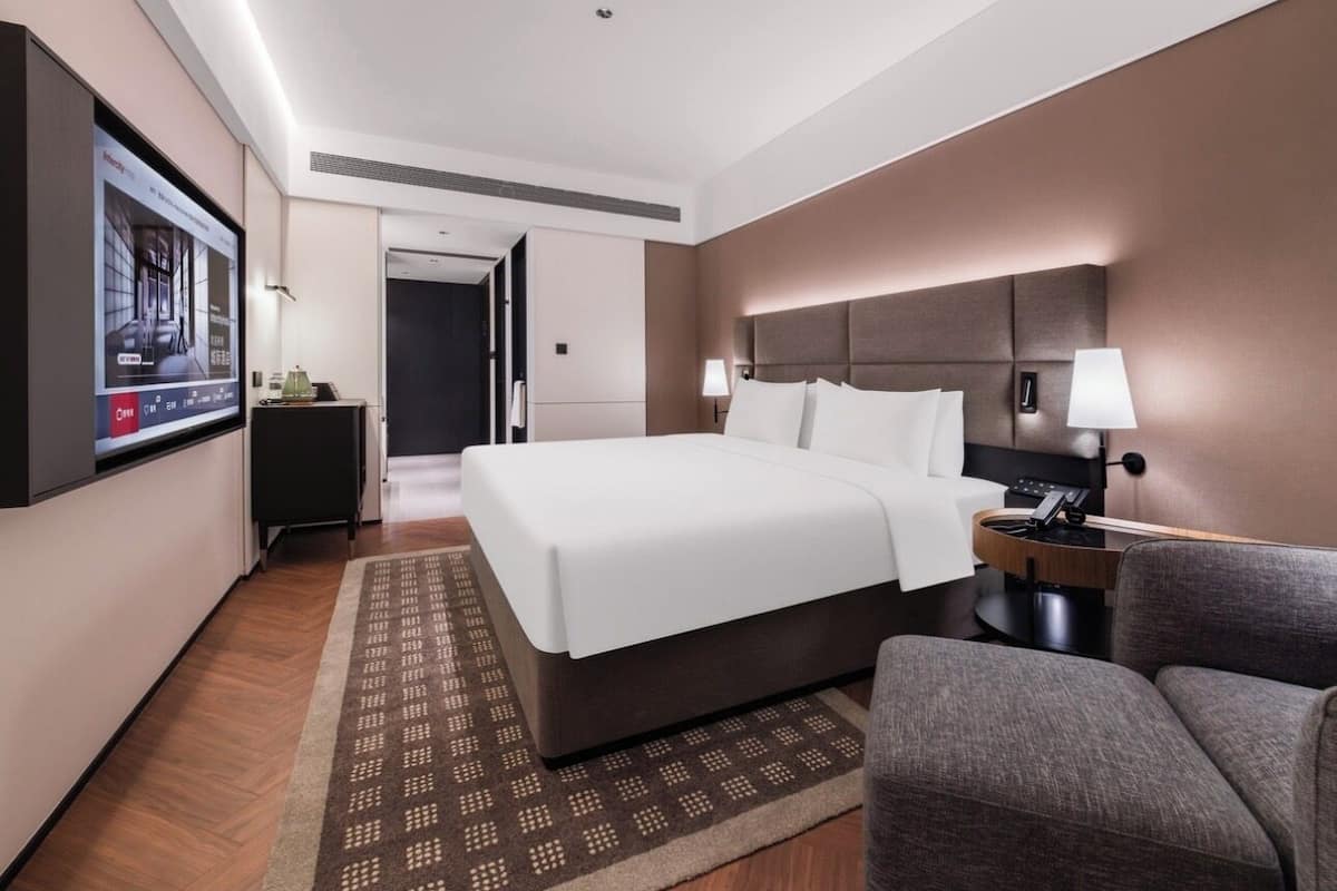 Quarto de hotel com uma cama grande, escrivaninha com luminária, televisão e um sofá.