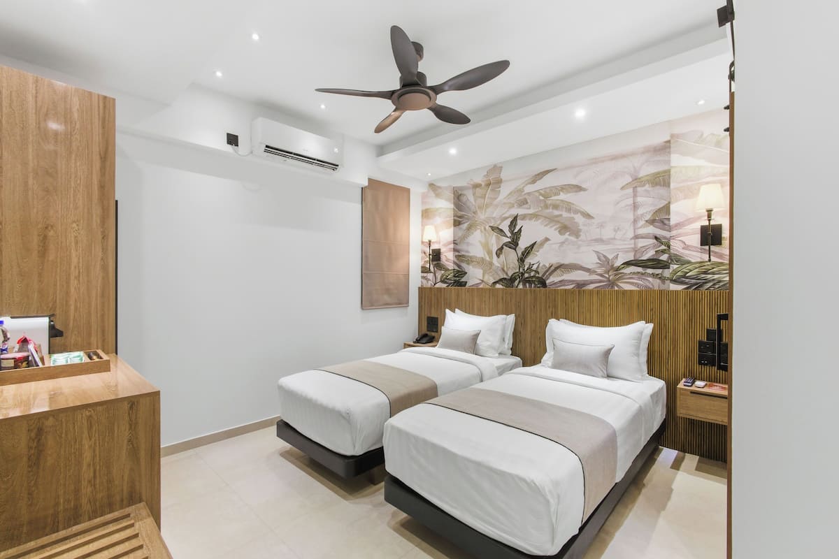 Quarto de hotel com duas camas, uma escrivaninha, um ventilador de teto, telefone fixo na parede e um mural com tema tropical.
