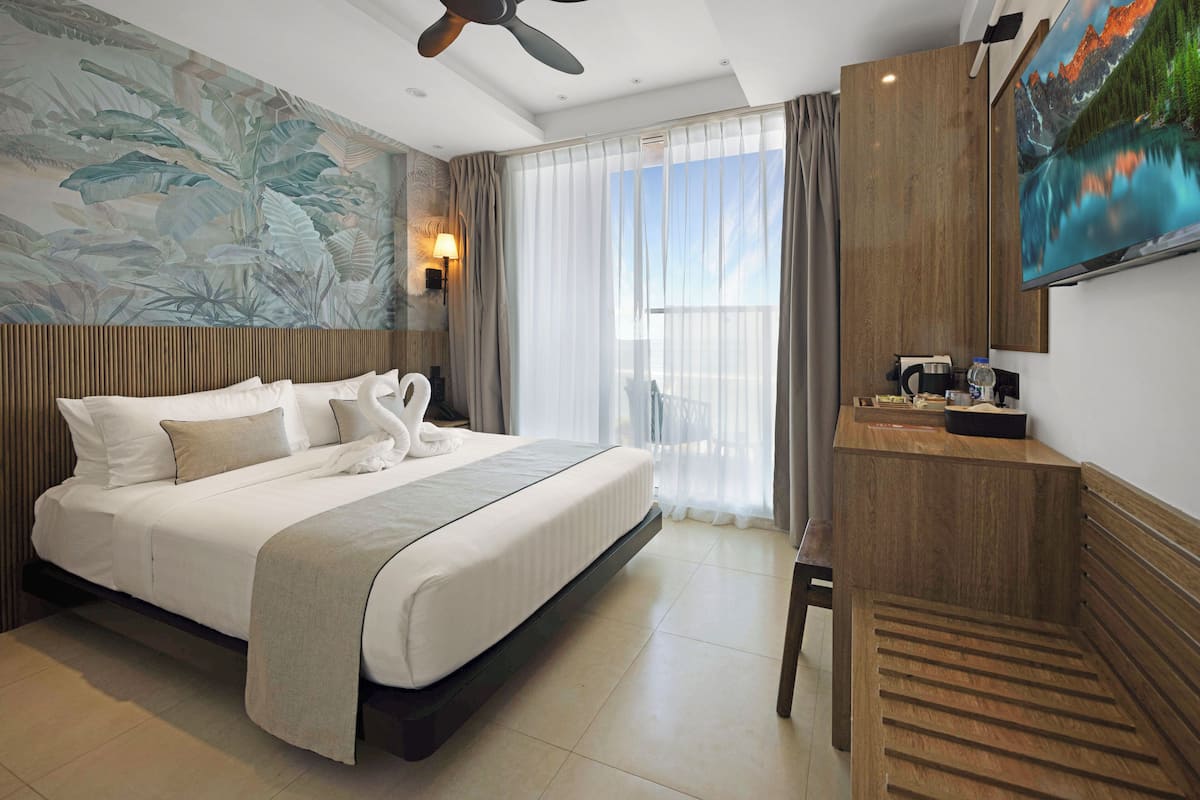 Quarto de hotel com cama, escrivaninha, ventoinha no teto, pintura e vista para o exterior.