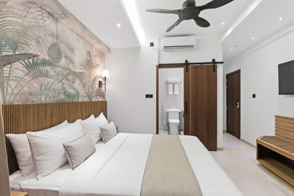 Quarto de hotel moderno com uma cama grande, um ventilador de teto, um aparelho de ar condicionado, uma cabeceira de madeira e uma televisão presa à parede.
