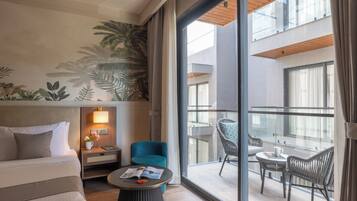 Quarto de hotel com varanda, cama, mesa de cabeceira, uma mesa com revista e uma cadeira.
