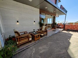 Exterior de um hotel com área de estar ao ar livre, vista clara do interior e uma placa com o nome do hotel.