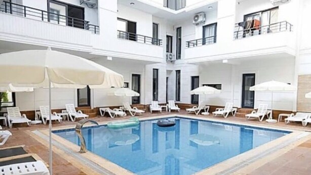 Uma piscina com espreguiçadeiras e guarda-sóis, rodeada por um edifício de vários níveis com varandas.
