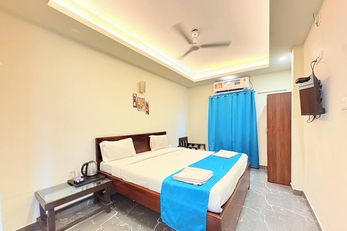Quarto de hotel com cama, escrivaninha, cadeira, ventilador de teto, televisão e guarda-roupa.