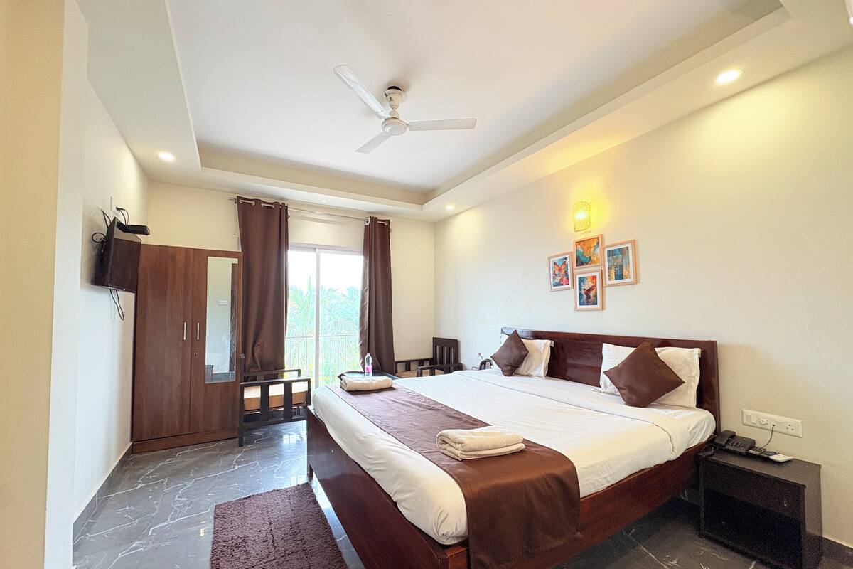 Quarto de hotel com cama, ventilador de teto, televisão, mesa de cabeceira e vista para o exterior.